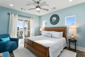 Quarto com cama, ventilador de teto, janela com cortinas, vista para o exterior e uma mesa de cabeceira com abajur.