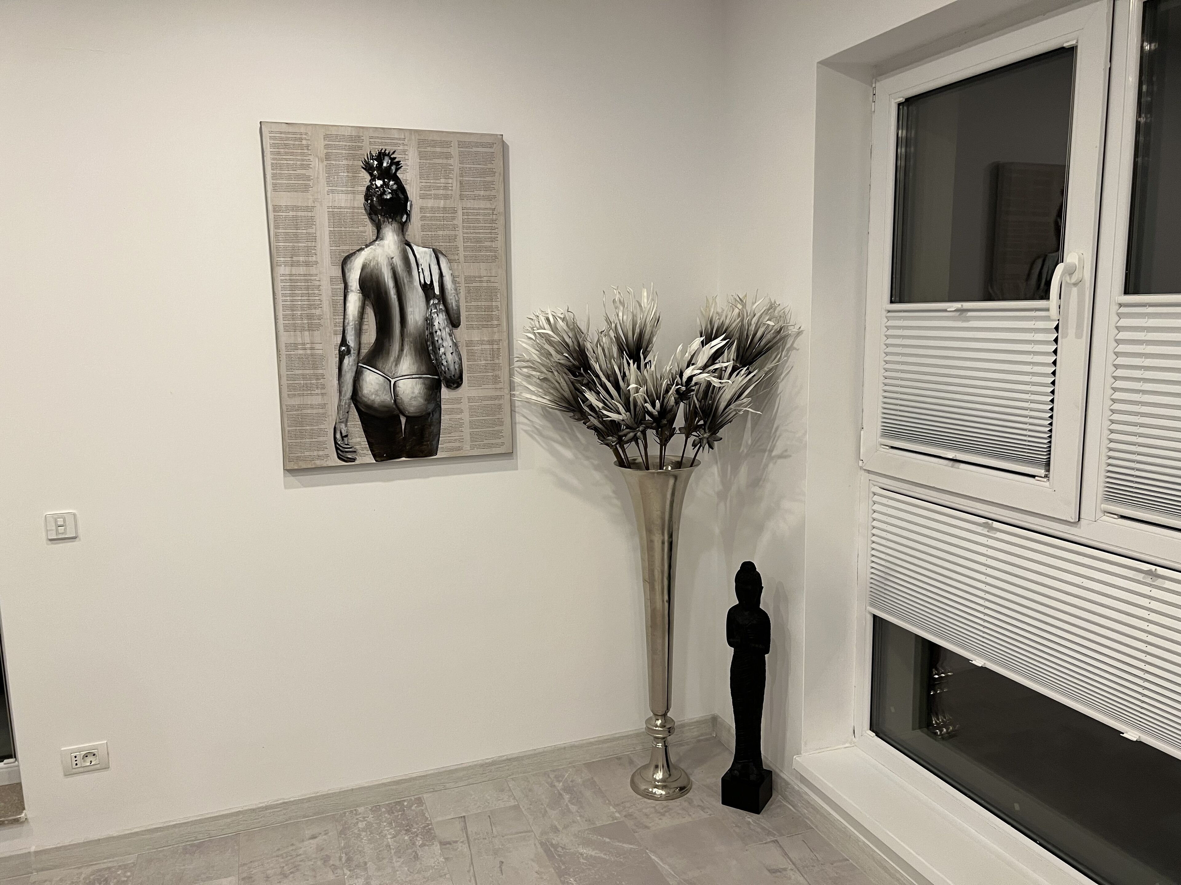 Um corredor com uma obra de arte em preto e branco emoldurada retratando uma mulher, um vaso alto com flores secas e uma estátua preta.