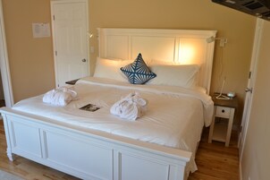 Cama bem arrumada com lençóis brancos e um travesseiro decorativo, mesa de cabeceira com abajur e porta com um aviso na parede.
