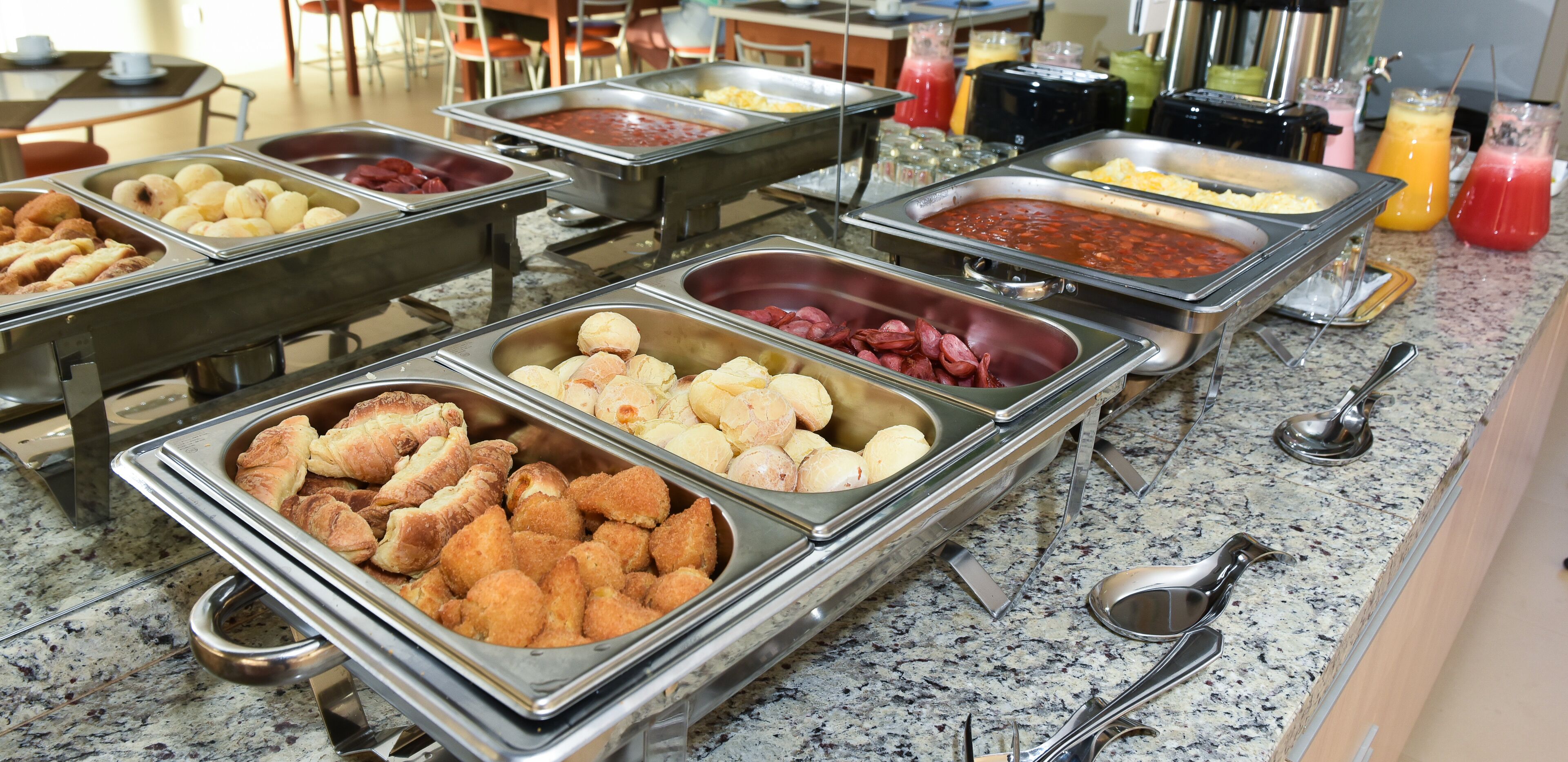 Buffet com diversos alimentos em recipientes de metal, incluindo pão, doces e diferentes tipos de carne.