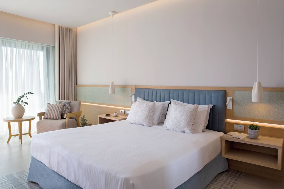 Quarto de hotel moderno com uma cama grande, mesas de cabeceira, uma cadeira e uma pequena mesa com uma planta.