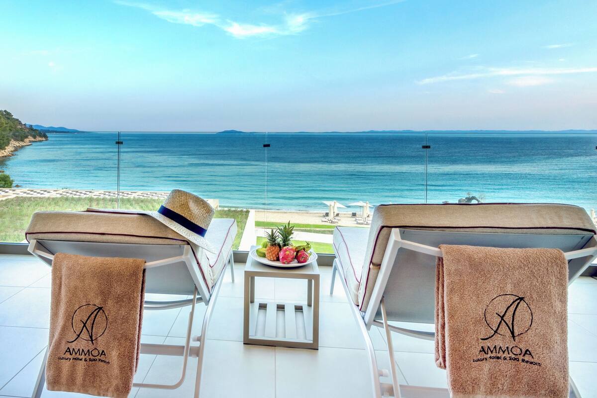 Varanda com duas poltronas de descanso, cada uma com uma toalha com o logotipo "AMMOA". Uma pequena mesa com frutas e um chapéu fica entre as poltronas. A vista inclui uma praia e o mar.