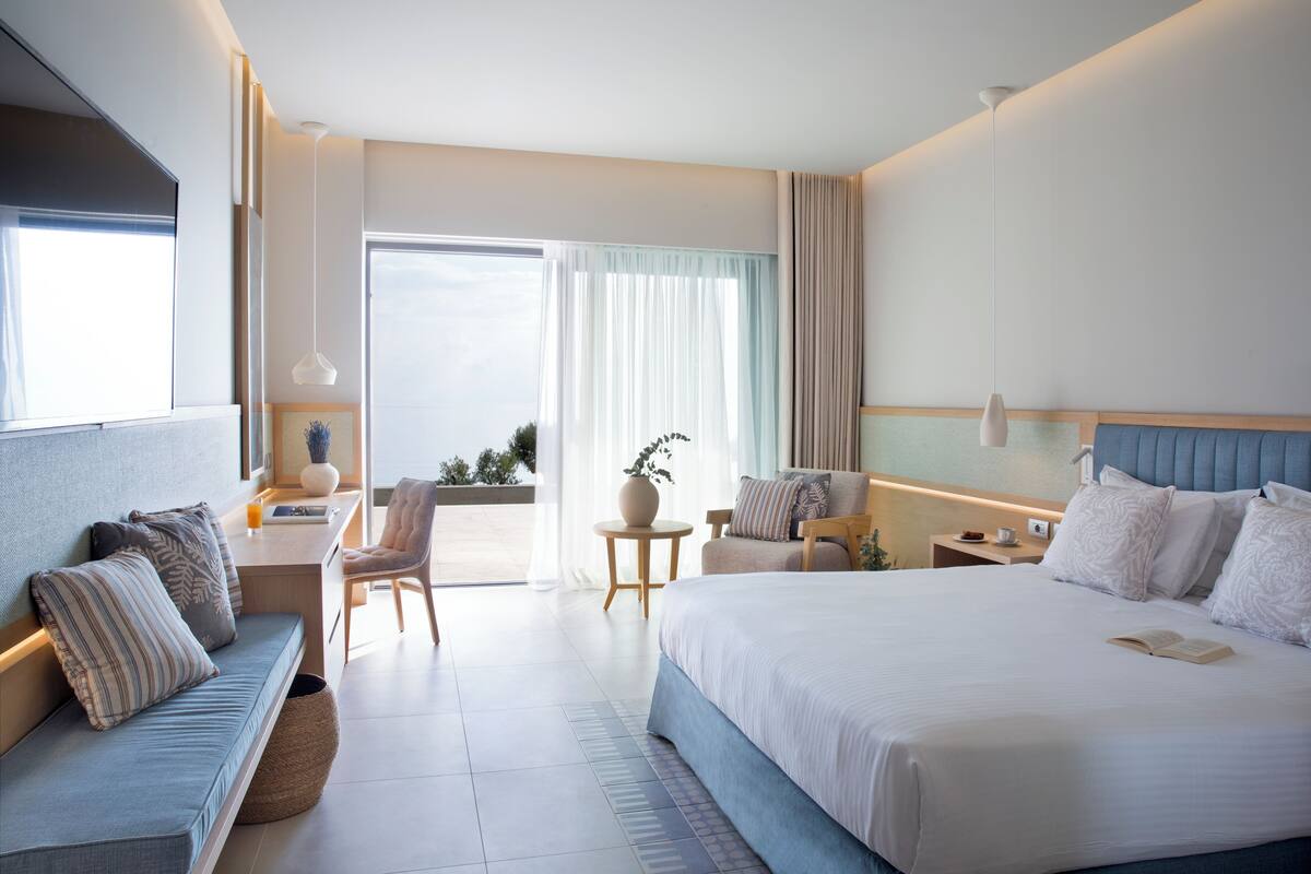 Quarto de hotel moderno com uma cama grande, área de estar com sofá e poltronas, uma pequena mesa com abajur, e vista para o exterior através de uma porta de vidro.