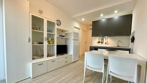Uma cozinha compacta com armários brancos, uma área de refeições com duas cadeiras e uma TV embutida.