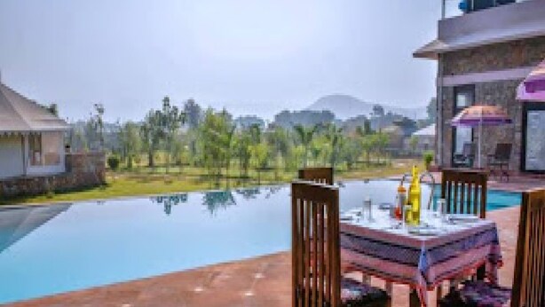 Uma área de piscina com mesa posta para refeições, rodeada de cadeiras, e vista para uma paisagem com árvores e colinas ao fundo.