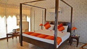Uma cama com dossel e estrutura de madeira, roupa de cama branca e detalhes em laranja. Há uma cadeira, uma pequena mesa com luminária e um ar-condicionado na parede.