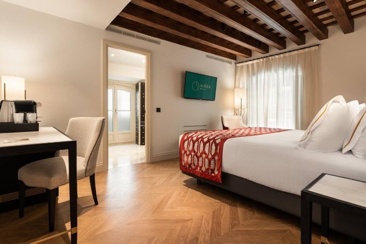 Quarto de hotel com uma cama, uma escrivaninha e uma cadeira. Há uma porta que dá para outro quarto.