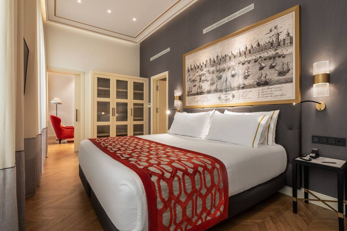 Quarto de hotel com uma cama grande, uma cadeira vermelha, um mapa histórico emoldurado e uma mesa de cabeceira com abajur.