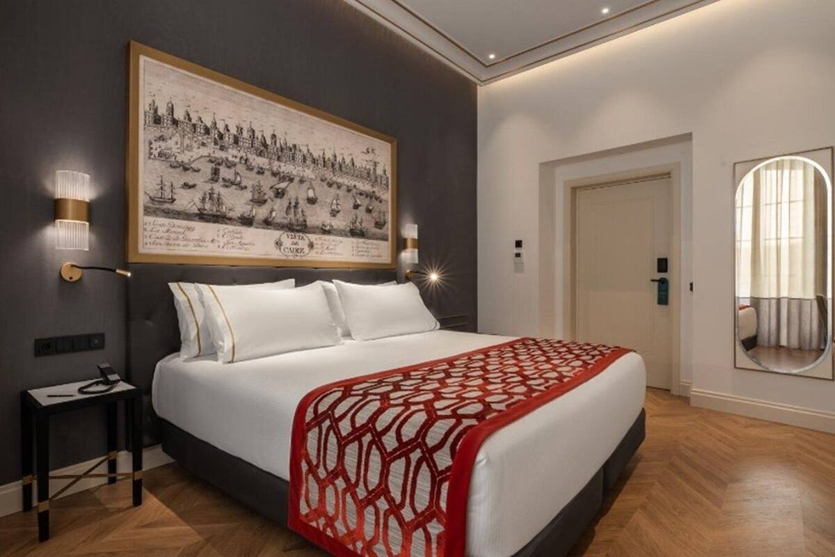 Quarto de hotel moderno com uma cama grande, um mapa histórico emoldurado na parede, uma mesinha de cabeceira com abajur e um espelho na parede.
