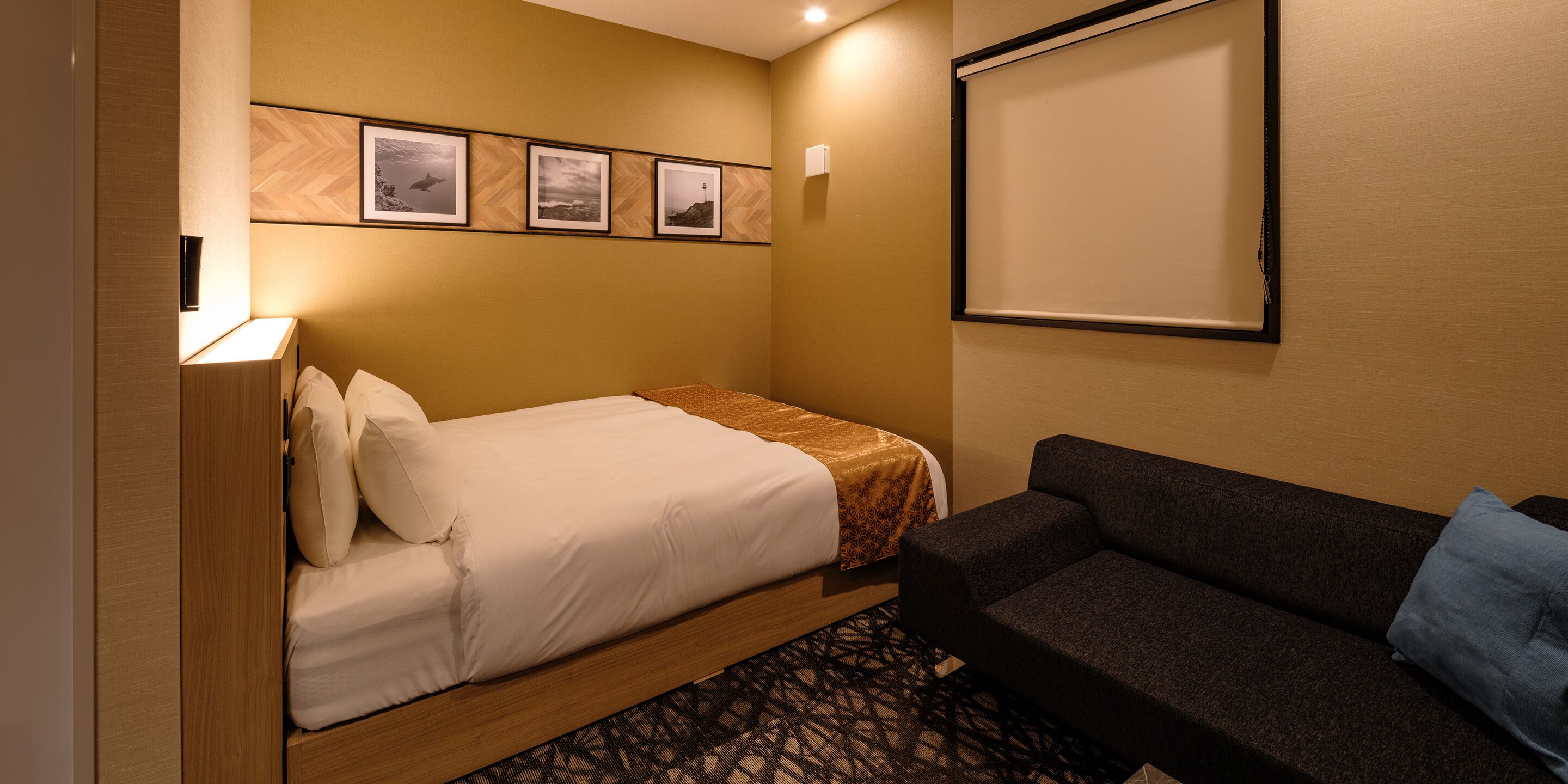 Quarto de hotel com uma cama, um sofá, uma tela de projetor fixada na parede e uma série de quadros pendurados.