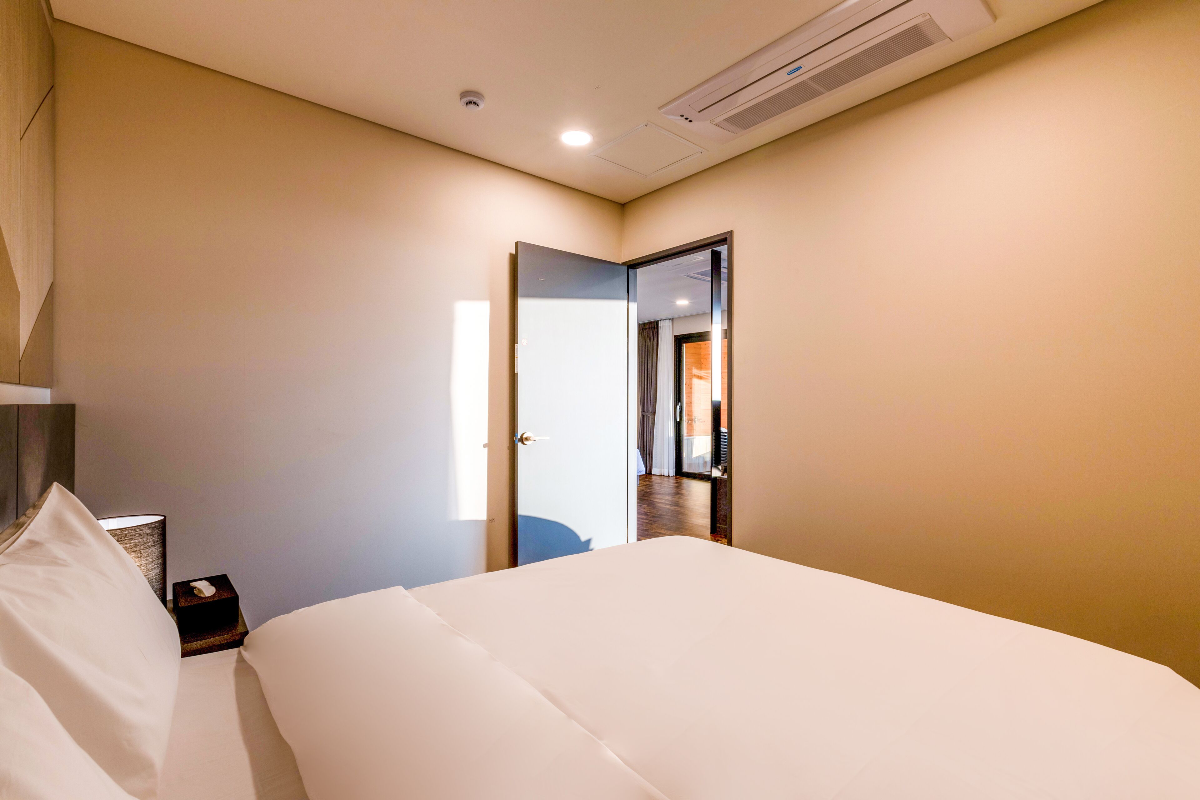 Quarto de hotel com uma cama grande, uma mesa de cabeceira com abajur e uma porta que dá para outro quarto.