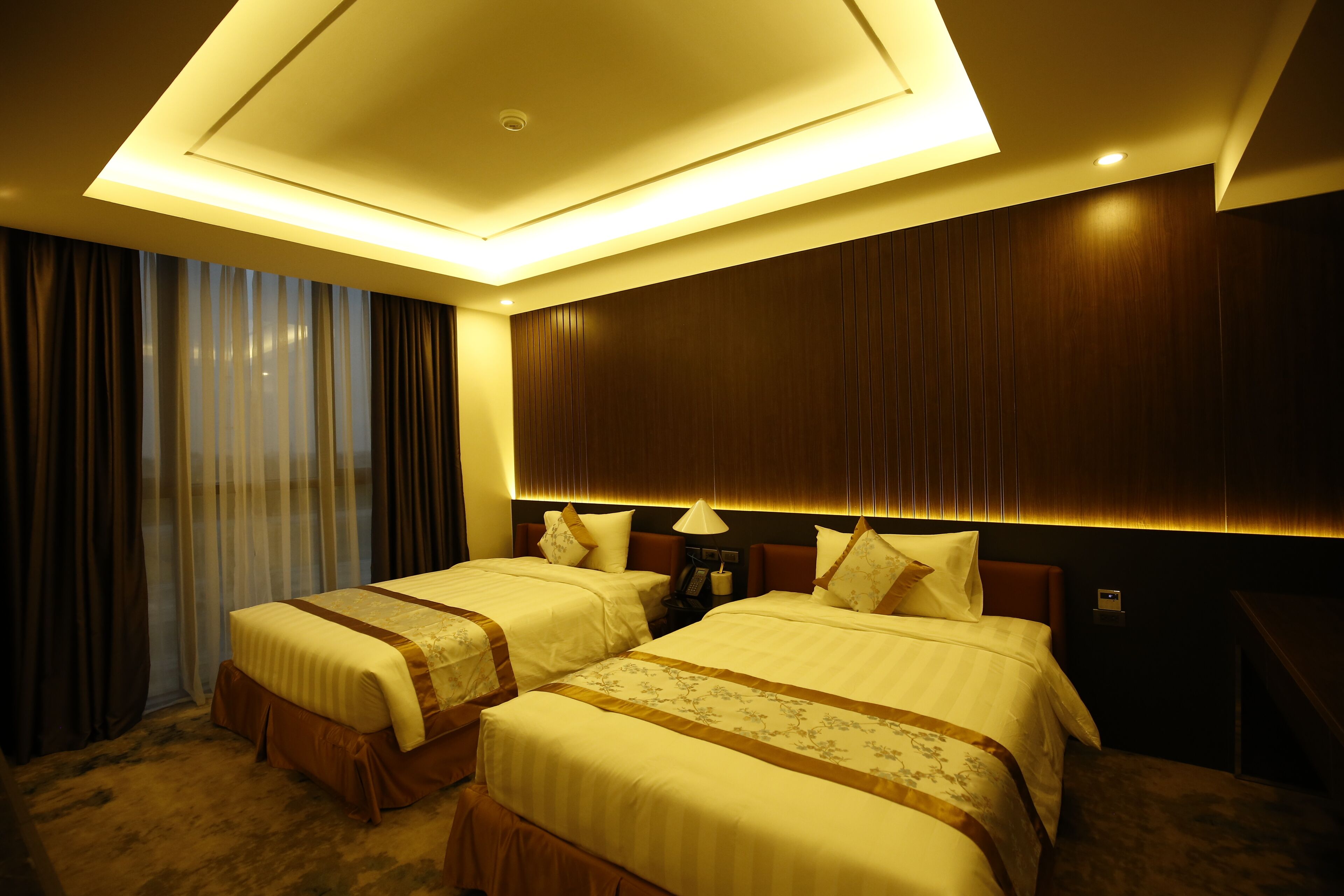Quarto de hotel com duas camas, uma janela grande com cortinas, uma luminária no teto e uma cabeceira de madeira.
