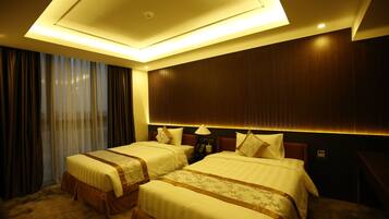 Quarto de hotel com duas camas, uma janela grande com cortinas, uma luminária no teto e uma cabeceira de madeira.