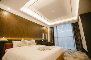 Quarto de hotel moderno com uma cama grande, cabeceira de madeira, escrivaninha e uma janela ampla com cortinas.