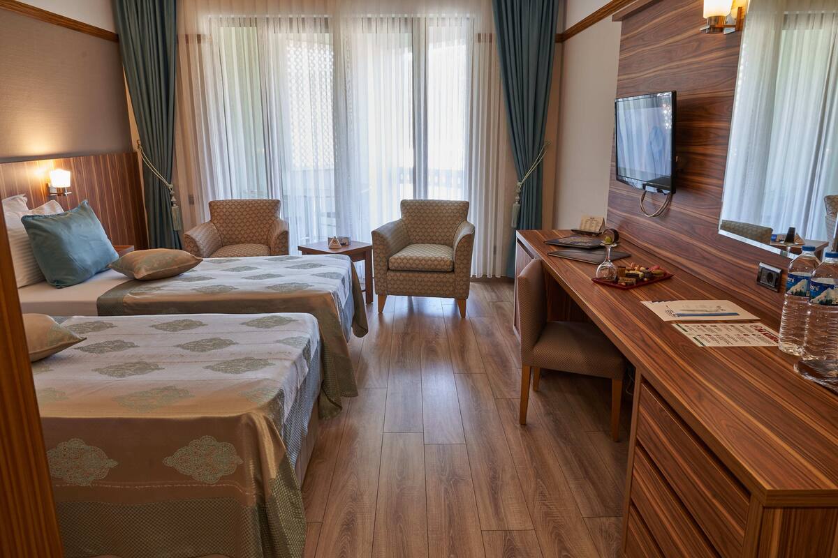 Quarto de hotel com duas camas, uma escrivaninha, uma cadeira, televisão e uma janela grande com cortinas.