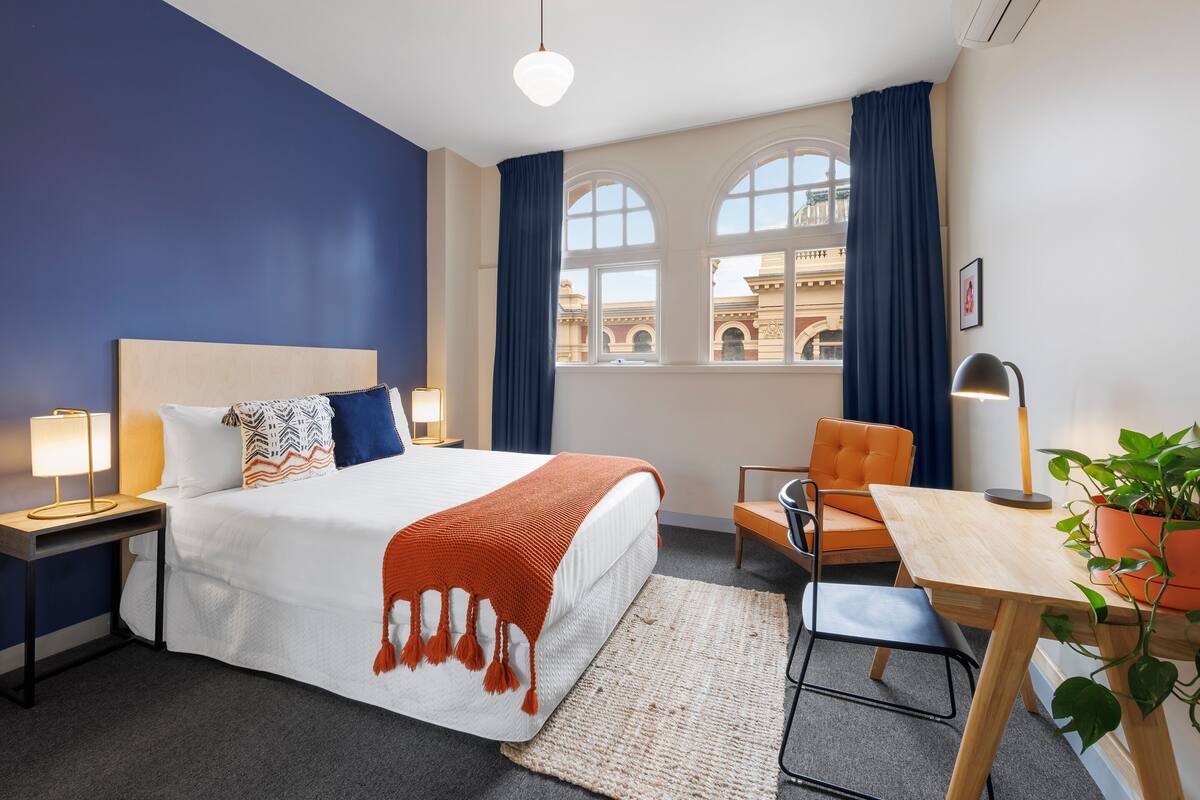Quarto de hotel com uma cama grande, duas mesas de cabeceira com abajures, uma escrivaninha com cadeira, uma planta em vaso e vista para um edifício pela janela.