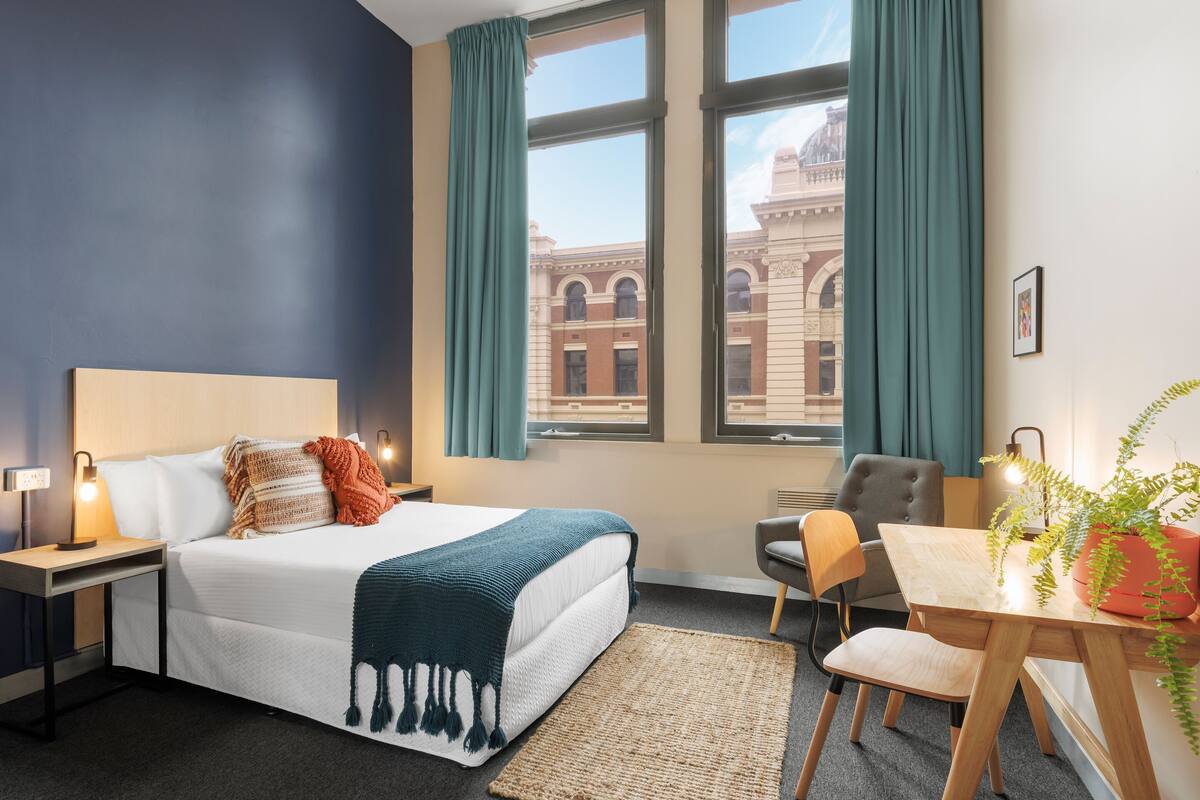 Quarto de hotel com cama, escrivaninha, cadeira, planta em vaso e vista de um edifício através de grandes janelas.