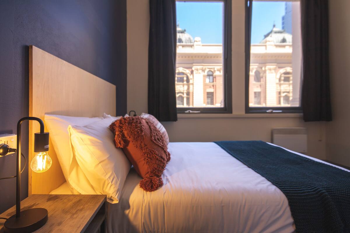 Quarto de hotel com cama, mesa de cabeceira com abajur e janela com vista para um edifício.