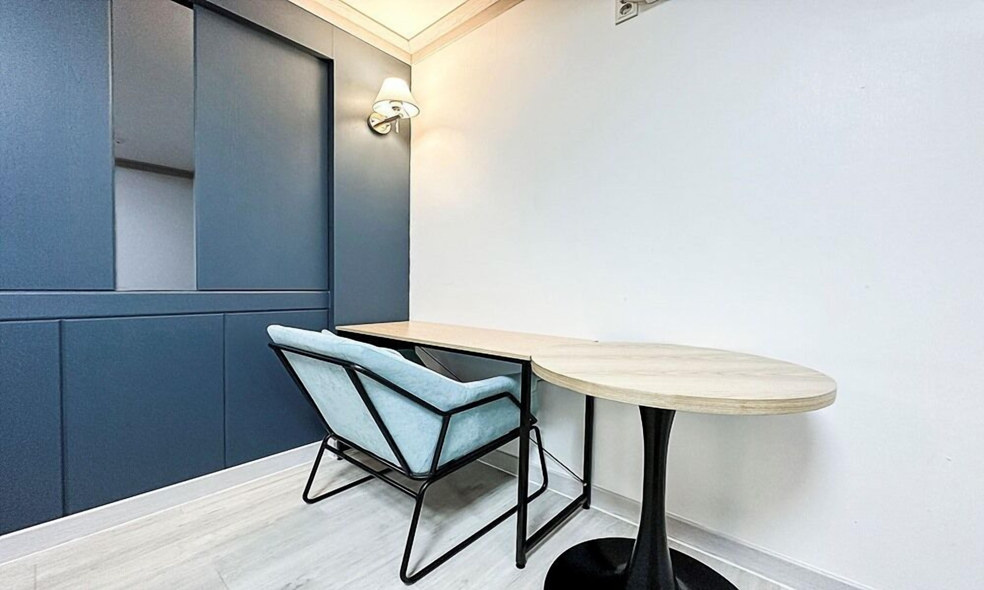 Quarto pequeno e moderno com mesa redonda e uma única cadeira, com acabamento em madeira clara e estrutura metálica preta.