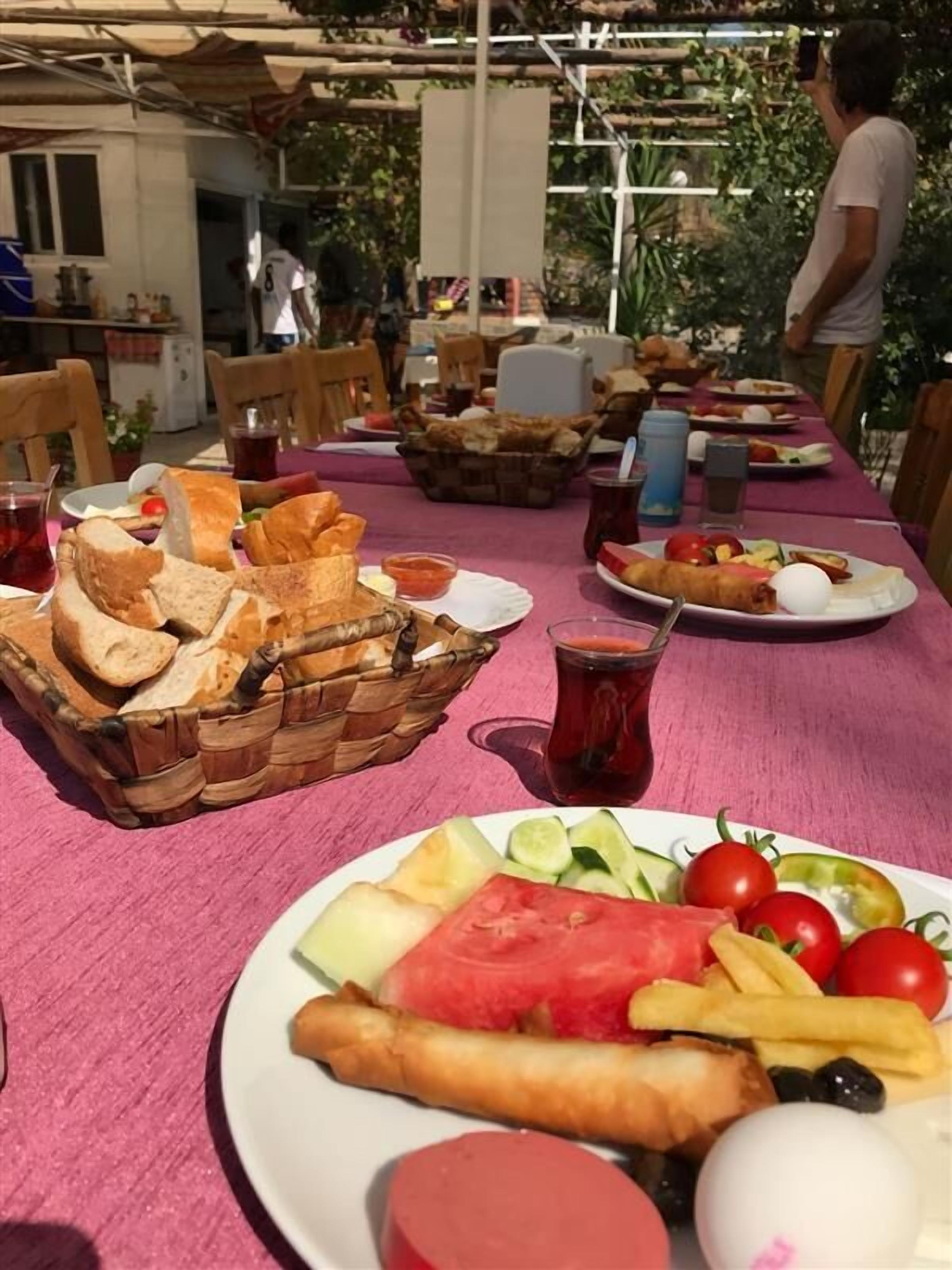 Mesa posta com diversos alimentos, incluindo pão, frutas e um prato com um item vermelho, possivelmente carne ou vegetal.