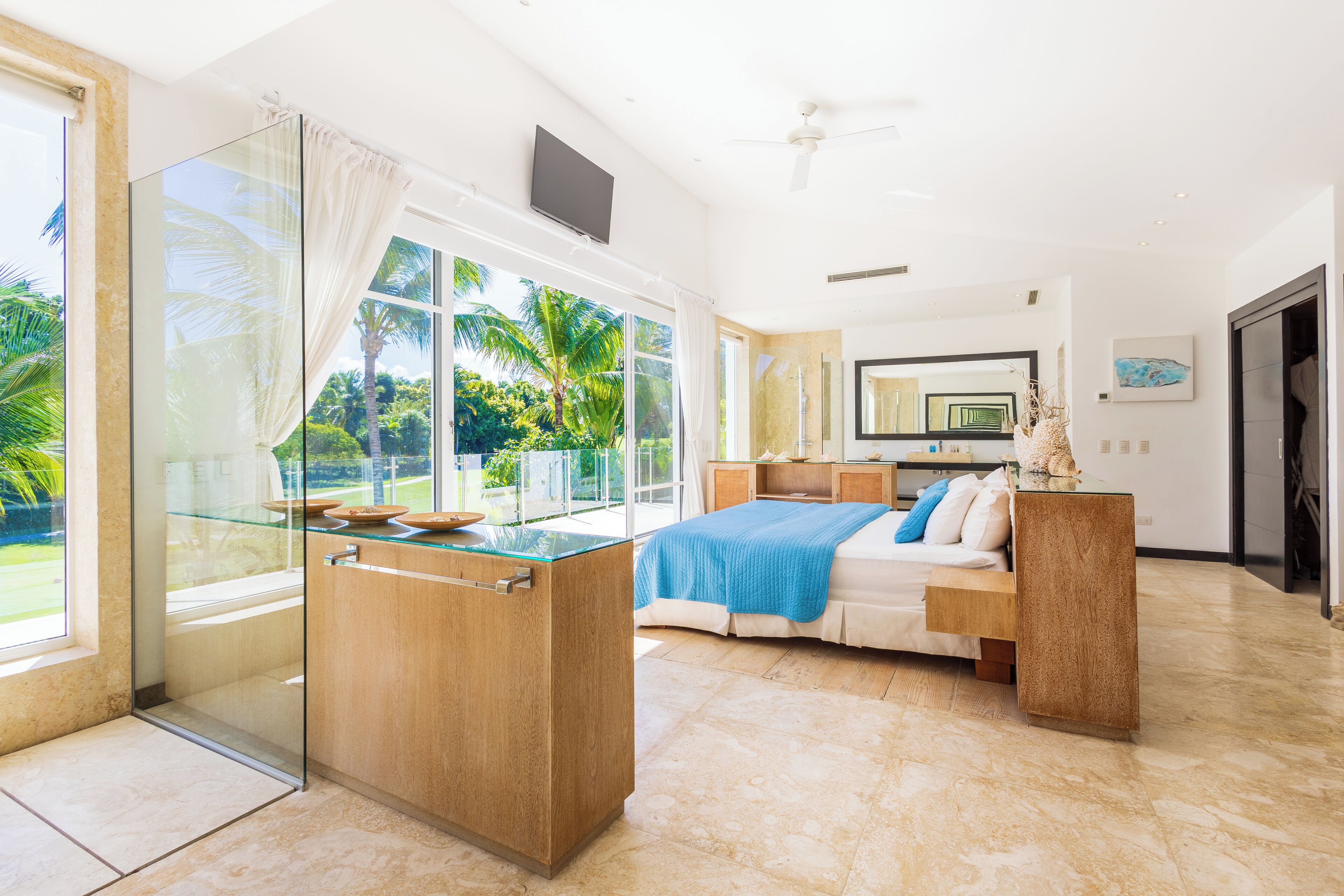 Quarto moderno com chuveiro de vidro, cama com manta azul e vista para palmeiras.