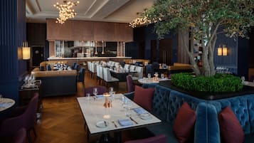 Restaurante com área de refeições central, bar e área de estar com árvore e sebe.