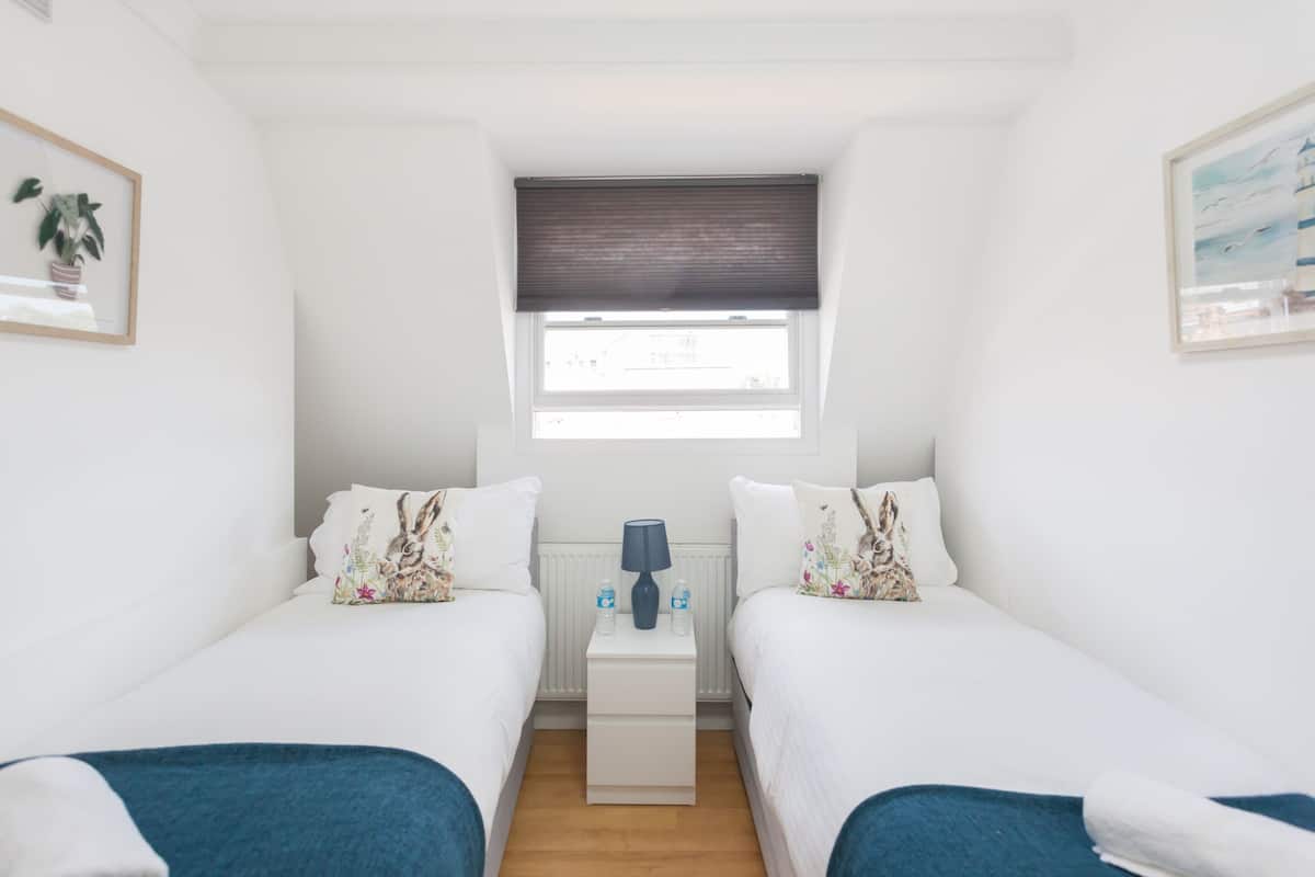 Quarto com duas camas de solteiro, uma pequena mesa de cabeceira com abajur e uma janela com cortina.