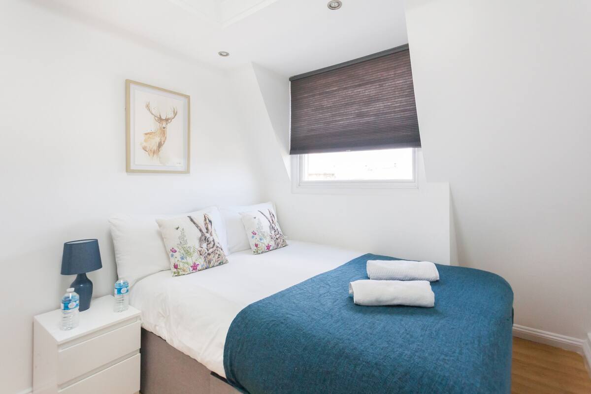 Quarto com cama, travesseiros, mesa de cabeceira com abajur e garrafas de água, quadro de veado e janela com cortina.