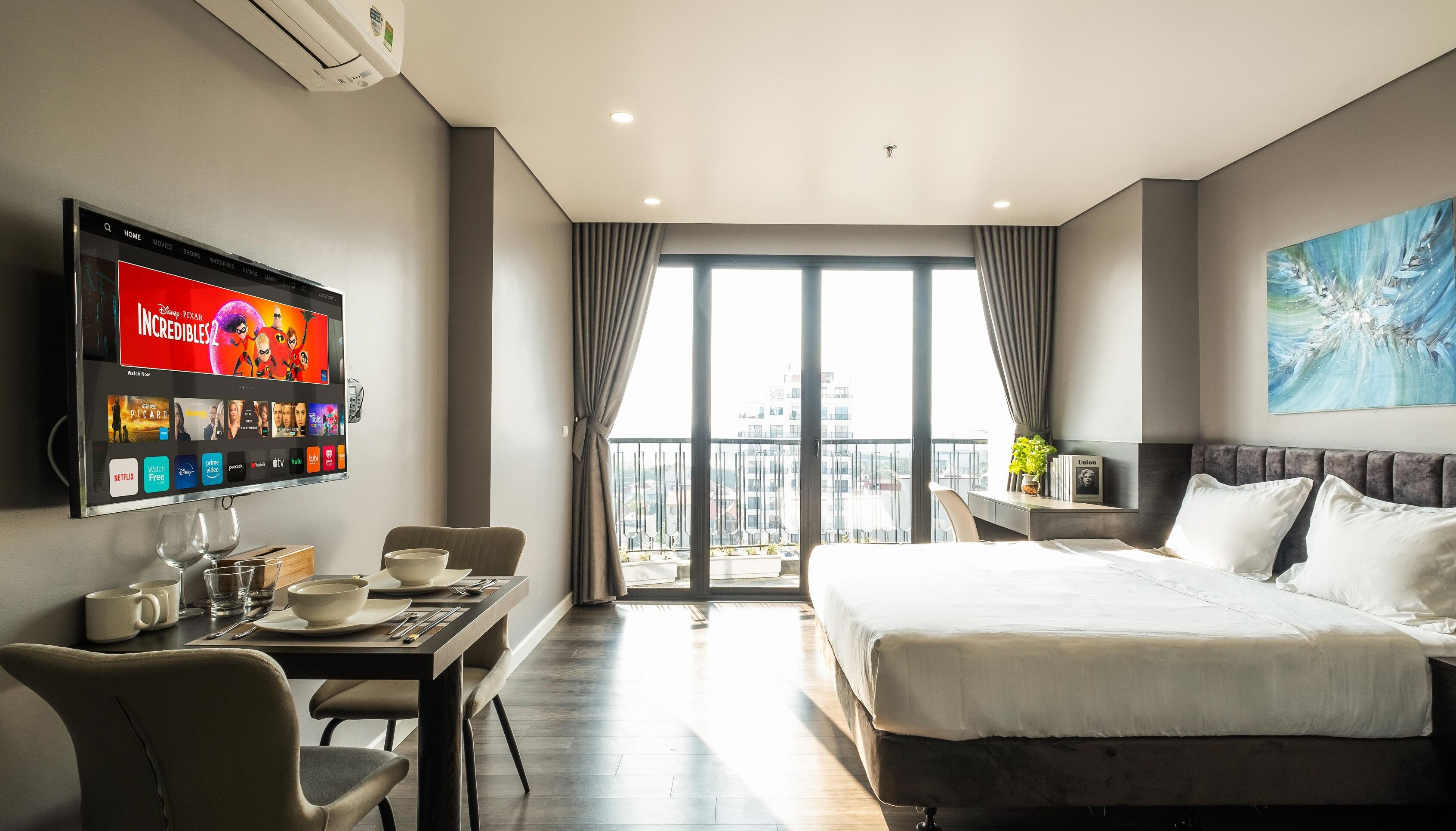 Quarto de hotel moderno com uma televisão grande exibindo um filme, uma varanda com cortinas, uma cama e uma escrivaninha com uma tigela e utensílios.