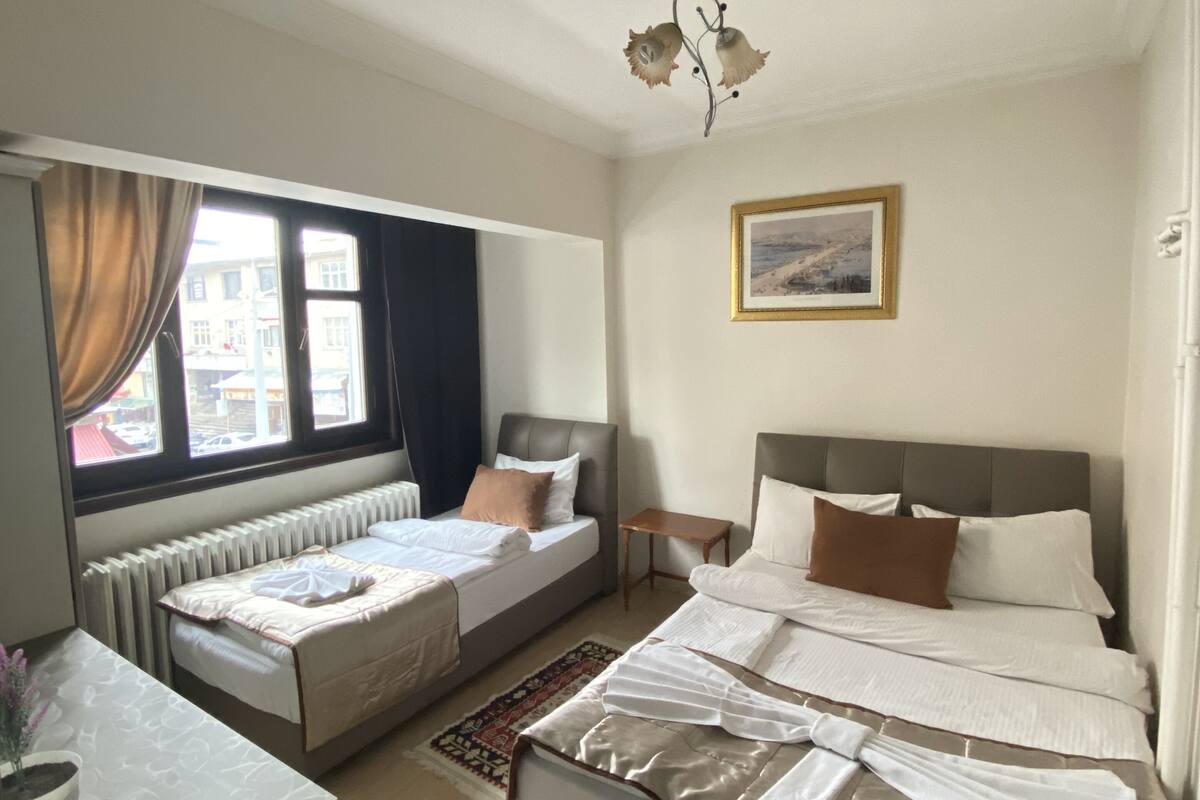 Quarto de hotel com duas camas, uma mesa de escritório com telefone, um radiador, uma janela com cortinas e um candeeiro de teto.