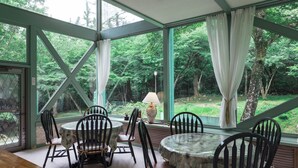 Uma área de refeições com mesa e cadeiras, janelas amplas com cortinas e vista para uma floresta.
