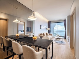 Quarto de hotel moderno com área de jantar, sofá, televisão e vista para o mar.