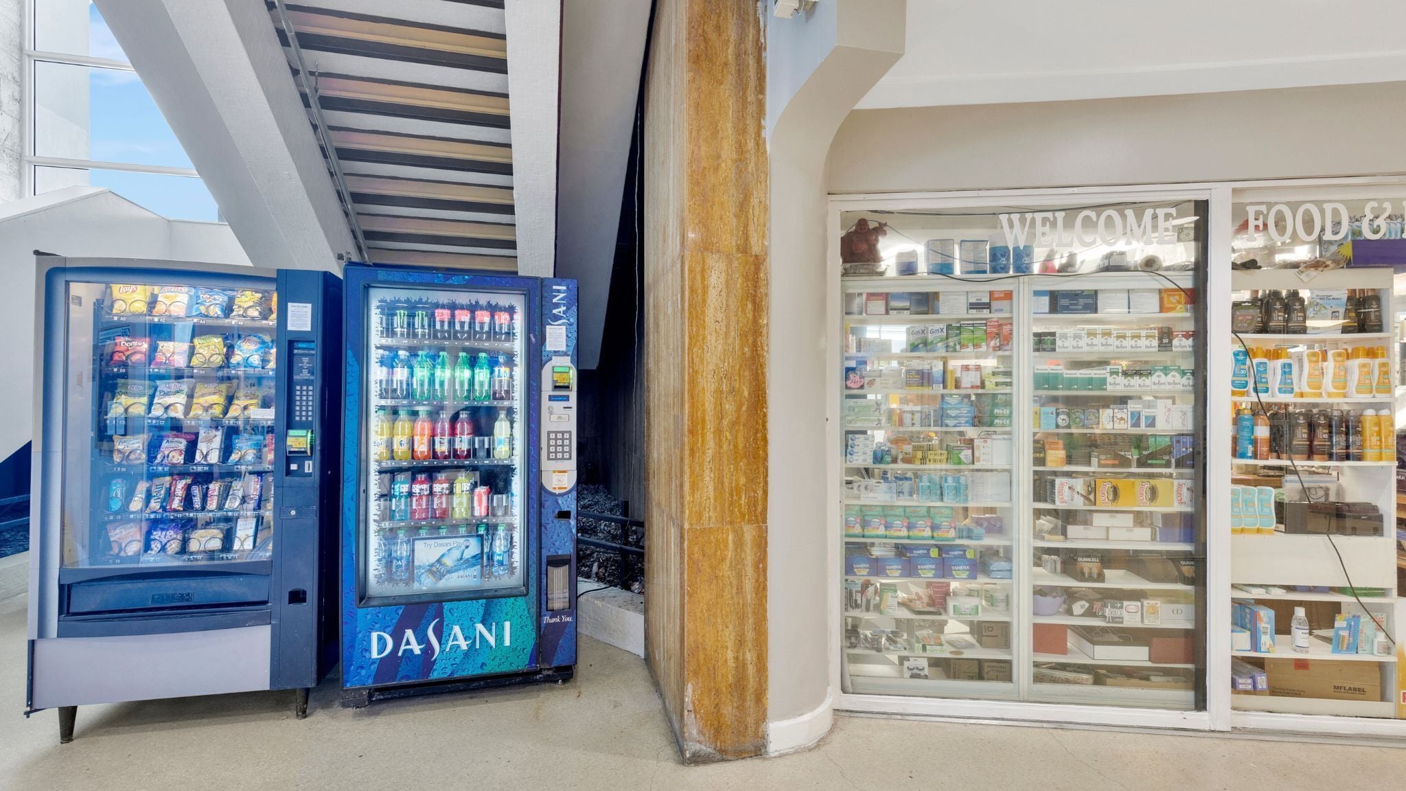 Uma máquina de venda automática com o logotipo DASANI, uma loja com um letreiro 'BEM-VINDO A ALIMENTOS' e um pilar grande.