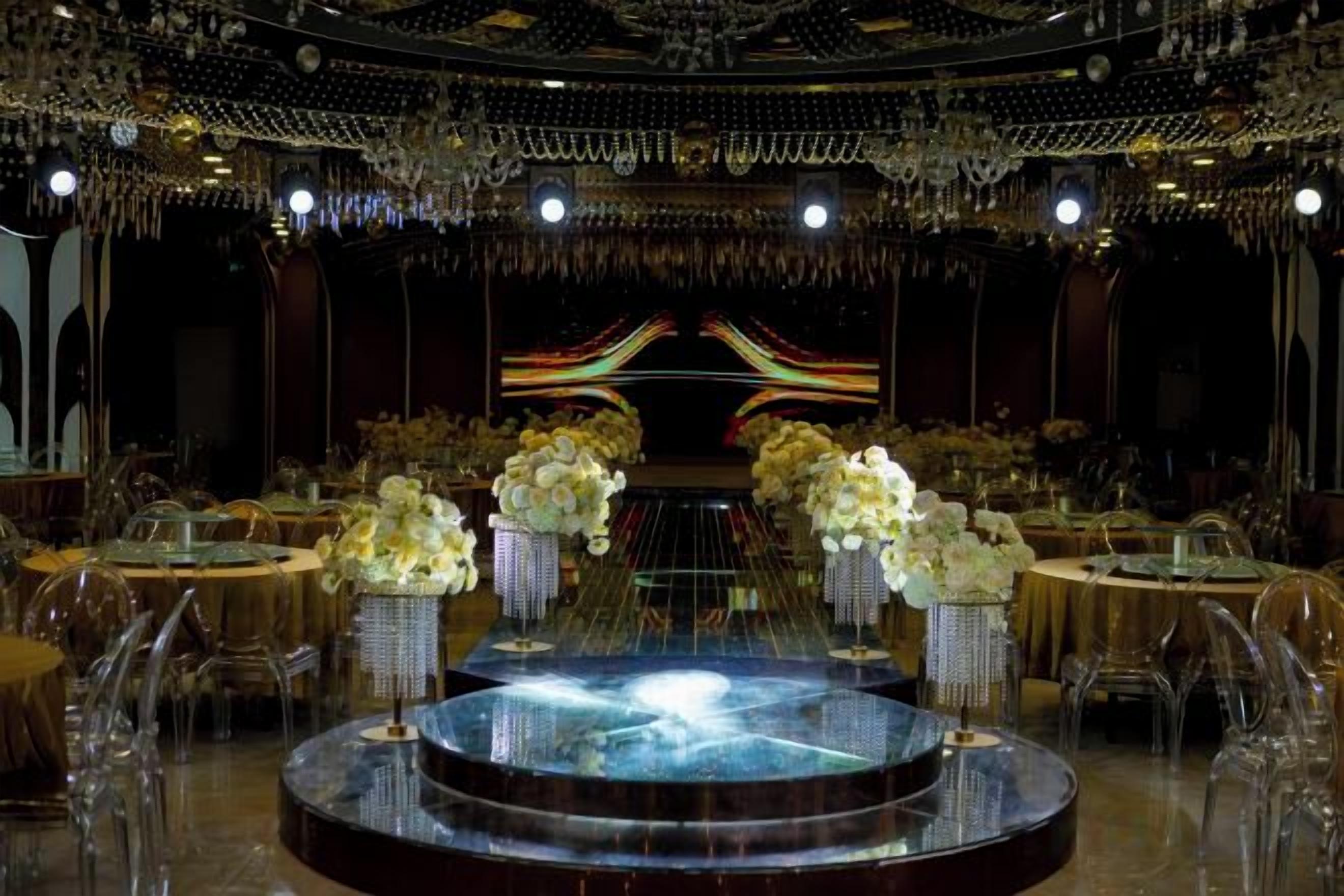 Uma grande sala de baile com palco central, mesas redondas com cadeiras e arranjos florais.