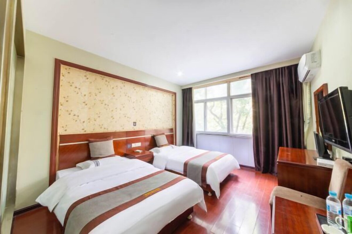 Quarto de hotel com duas camas, cabeceira de madeira, uma janela grande com cortinas, uma escrivaninha com cadeira e televisão de tela plana.