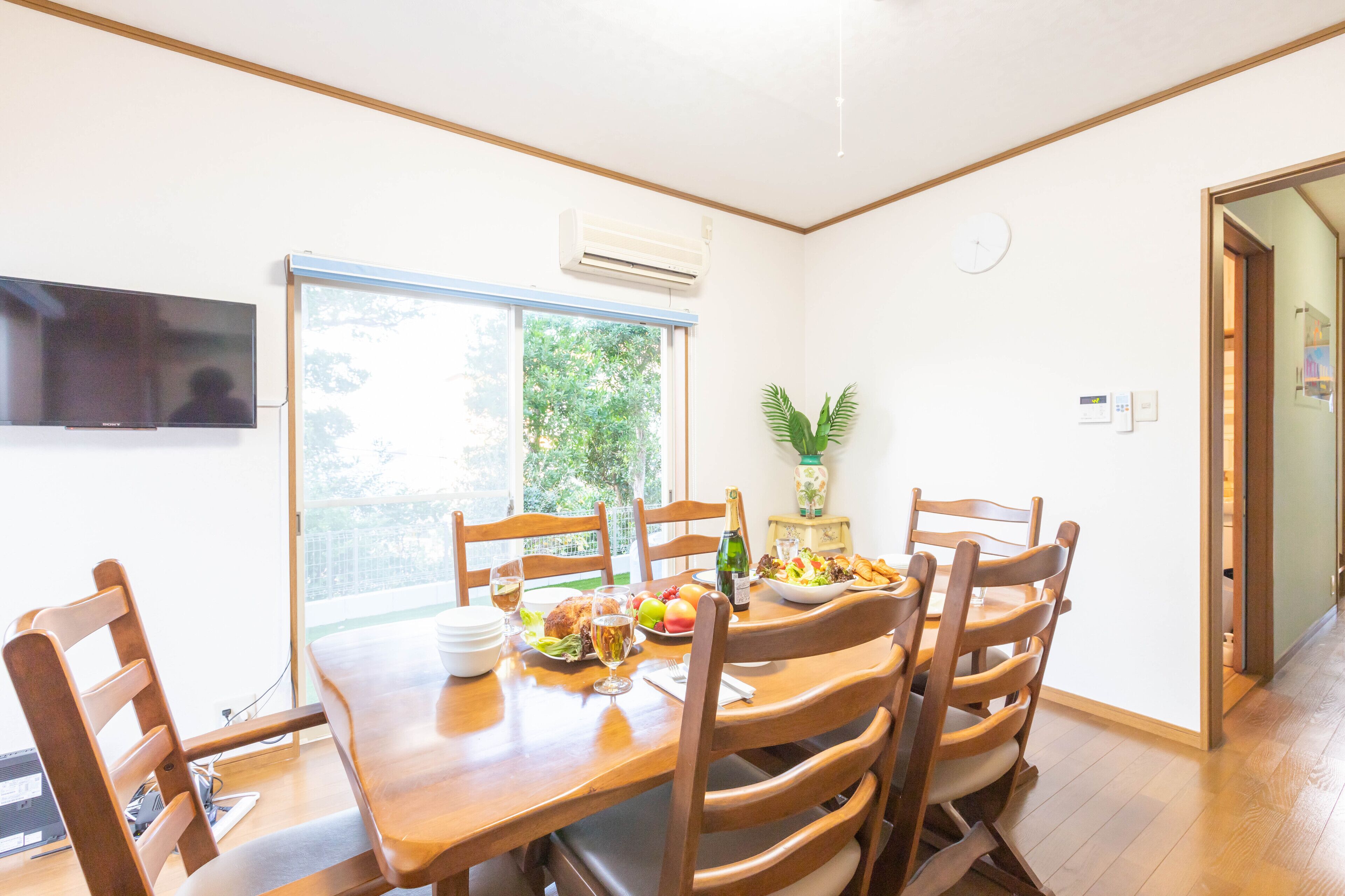 Uma sala de jantar com mesa de madeira, cadeiras, televisão e janela com vista para árvores.