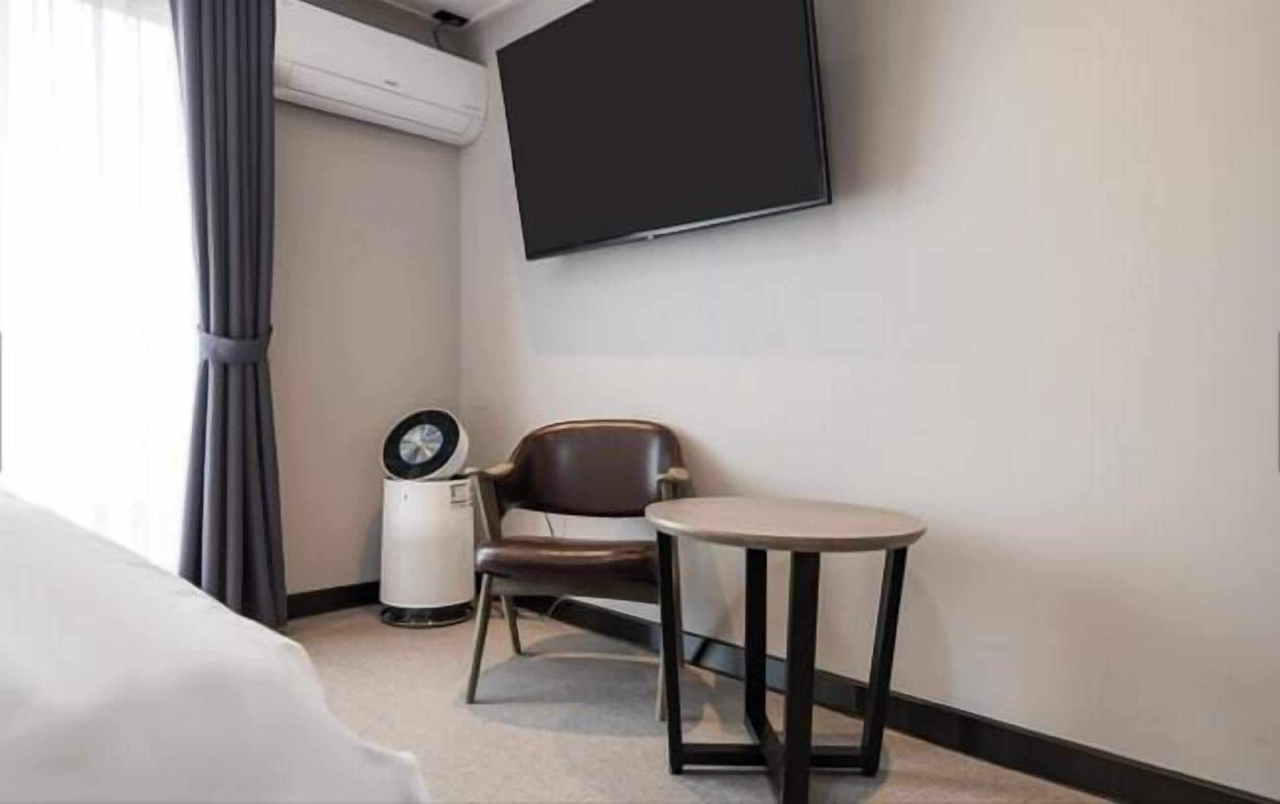 Quarto de hotel com cama, uma cadeira, uma mesinha, televisão fixada na parede e janela com cortinas.