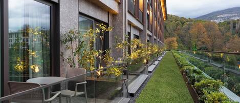 Varanda de hotel moderna com guarda-corpos de vidro, área de estar externa e vista para a cidade e colinas.