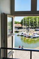 Vista da janela com vista para uma marina, barcos atracados, um calçadão pavimentado e um edifício ao fundo.