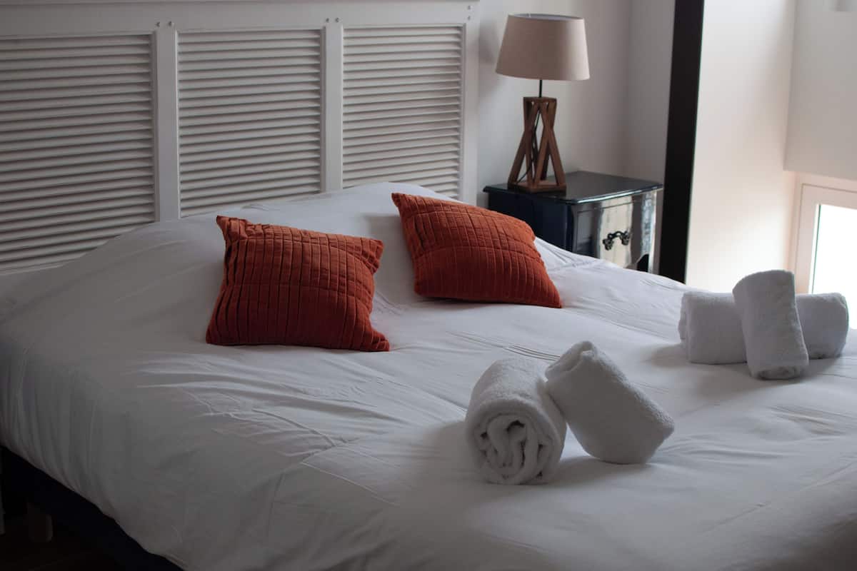 Cama bem arrumada com lençóis brancos, dois travesseiros laranjas e toalhas brancas dobradas em um quarto de hotel.