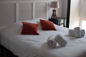 Cama bem arrumada com lençóis brancos, dois travesseiros laranjas e toalhas brancas dobradas em um quarto de hotel.