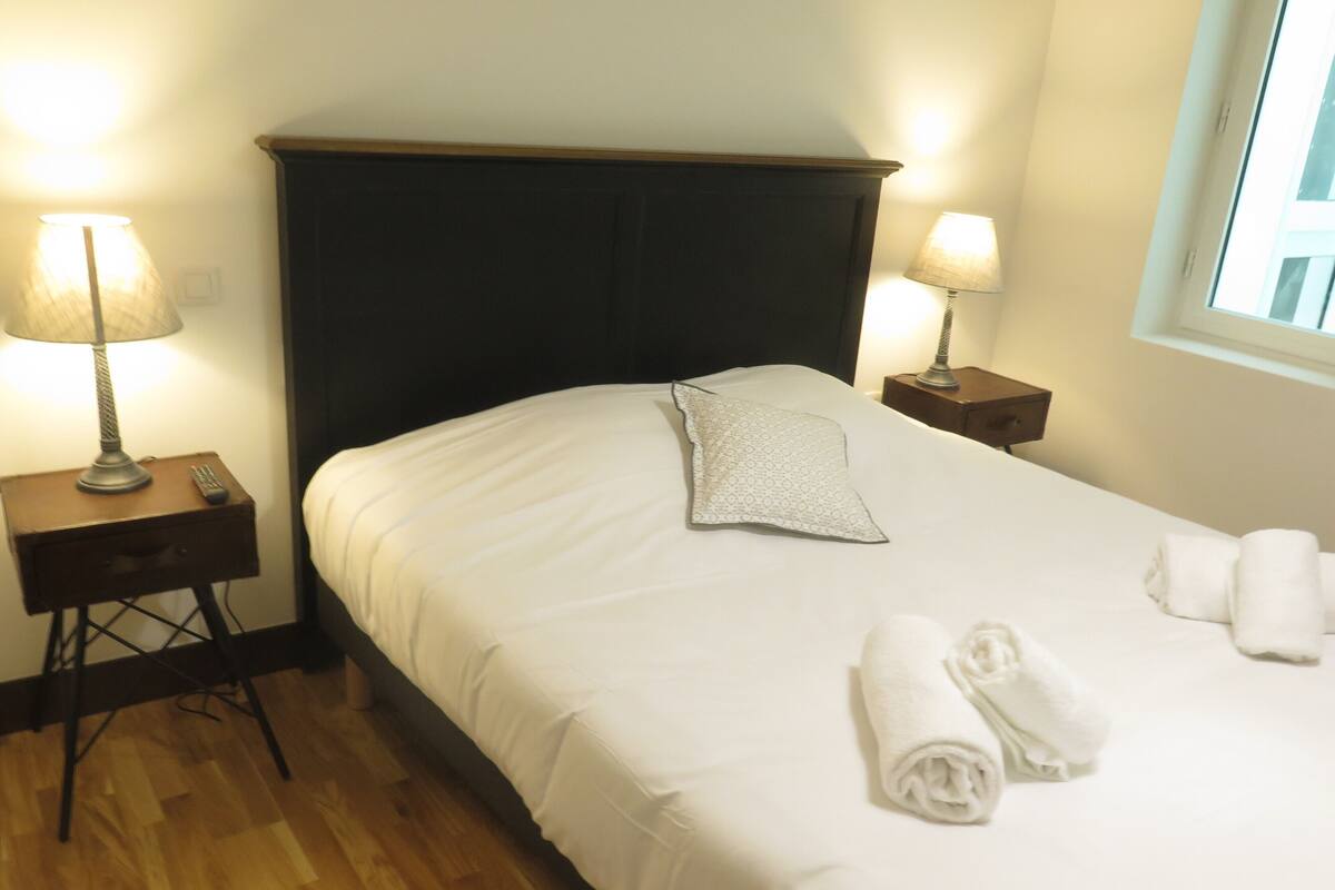 Cama bem arrumada com lençóis brancos, duas luminárias de cabeceira e uma janela com luz do dia.