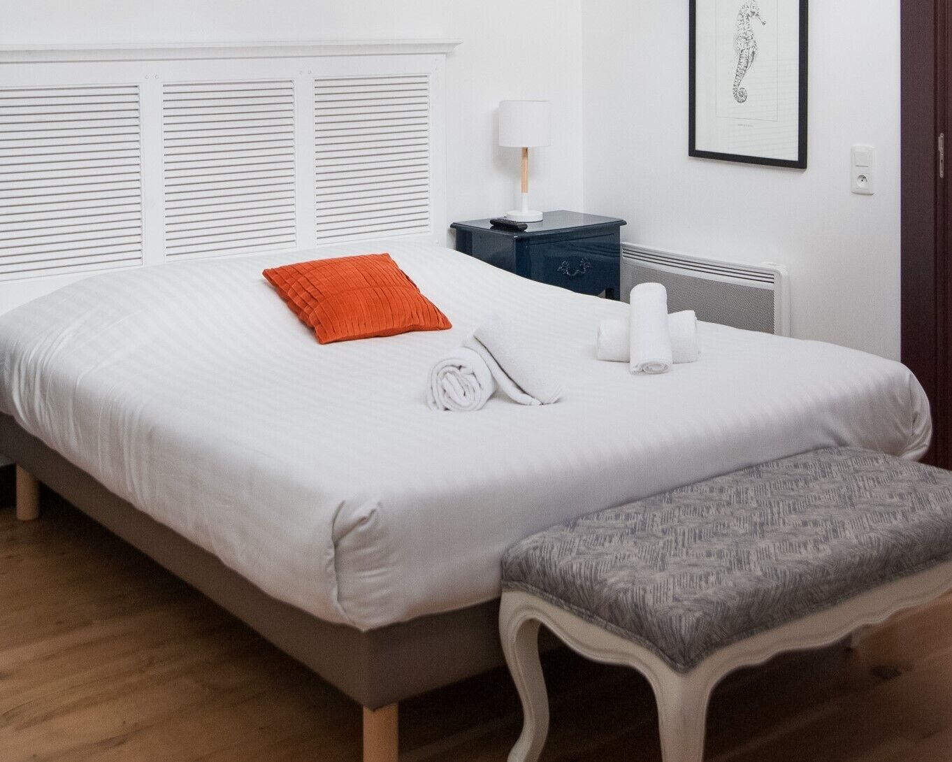 Cama bem arrumada com roupa de cama branca, um travesseiro laranja e toalhas dobradas. Uma pequena mesa de cabeceira com abajur e um quadro emoldurado fica ao lado da cama.