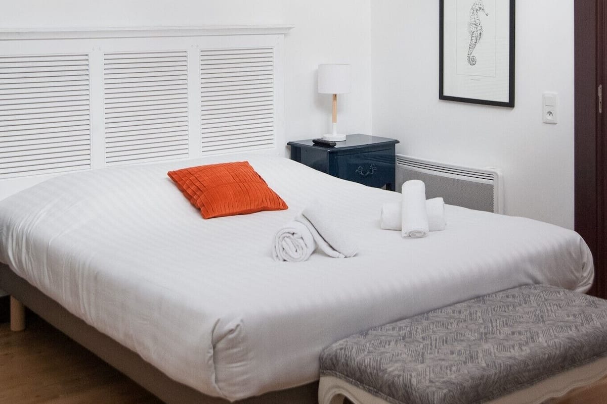 Cama bem arrumada com roupa de cama branca, um travesseiro laranja e toalhas dobradas. Uma pequena mesa de cabeceira com abajur e um quadro emoldurado fica ao lado da cama.