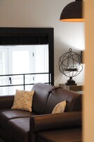 Um sofá de couro marrom com almofadas estampadas, uma janela com moldura preta e uma esfera de metal decorativa sobre uma mesa.