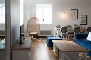 Uma sala de estar acolhedora com um sofá azul, uma mesa de centro de madeira, uma poltrona de vime e um espaço de jantar com mesa e cadeiras.