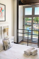 Quarto de hotel com cama, mesa de cabeceira, abajur, janela com vista para barcos e água, e um quadro na parede.