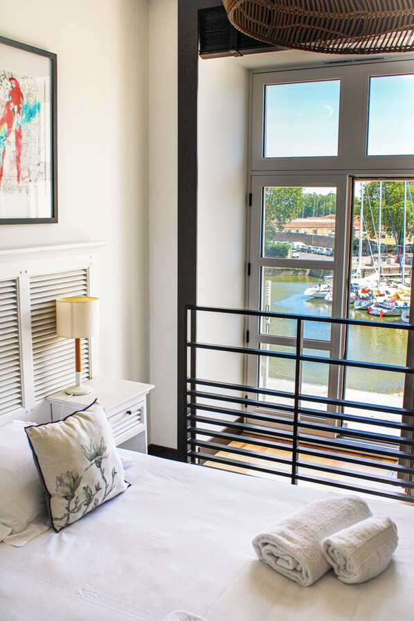 Quarto de hotel com cama, mesa de cabeceira, abajur, janela com vista para barcos e água, e um quadro na parede.