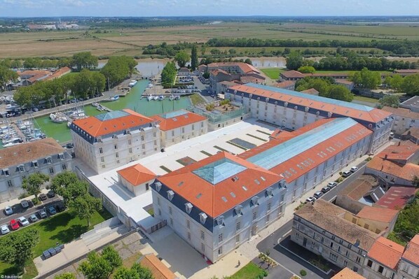 Um edifício grande e histórico, com várias alas e um pátio central, rodeado por uma marina e campos verdes.