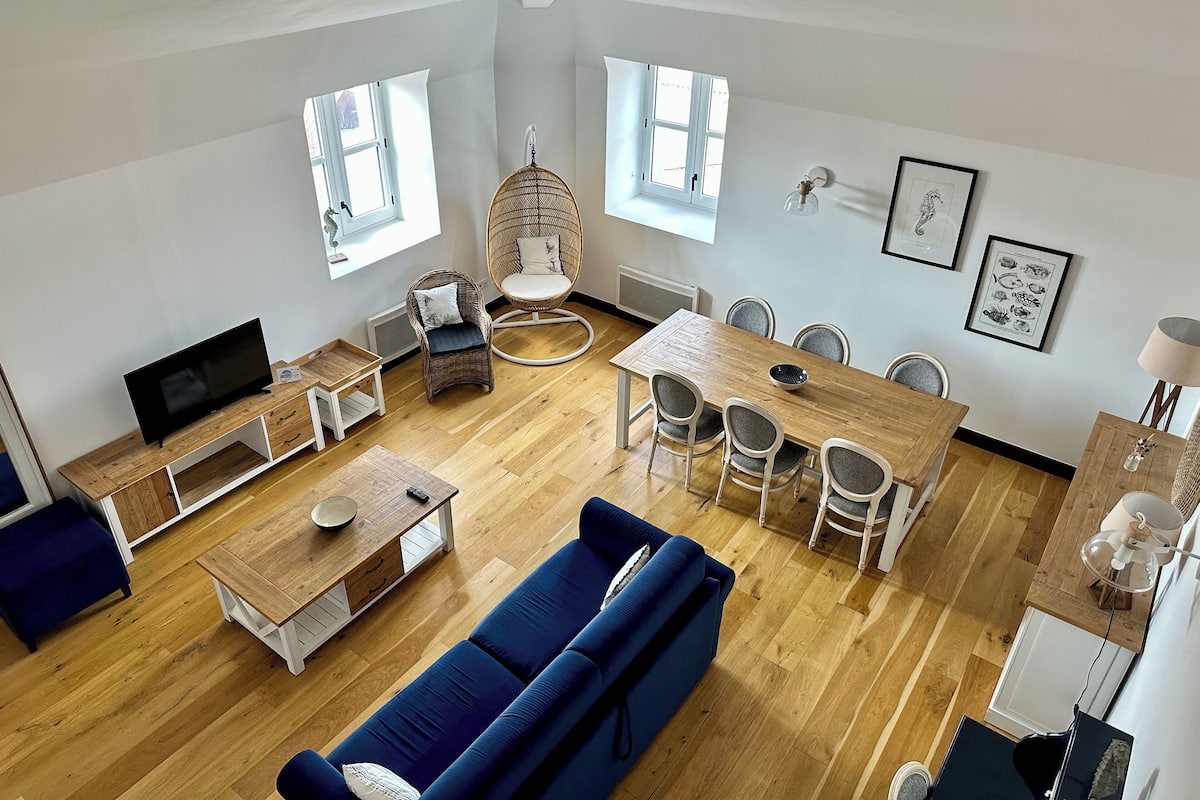 Uma sala de estar ampla com piso de madeira, um sofá azul, uma mesa de jantar com cadeiras, uma televisão e uma poltrona de vime.